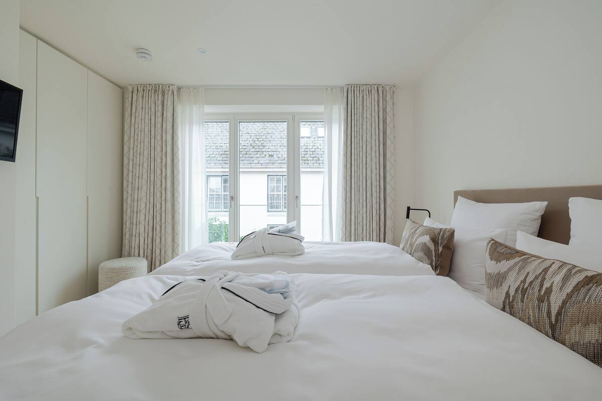 Quarto de hotel com duas camas, uma televisão de tela plana, uma janela com cortinas e vista para prédios.