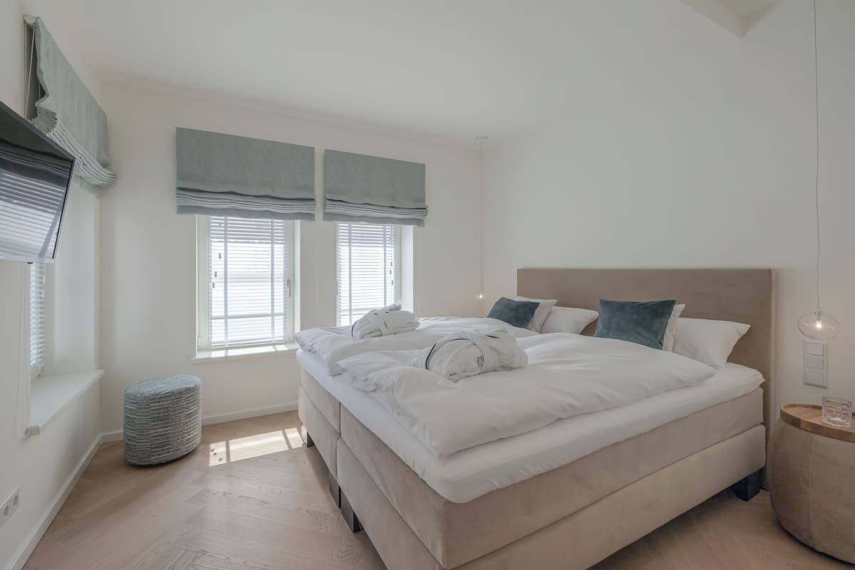 Quarto com cama, mesa de cabeceira, janela com persianas e um pequeno banquinho.