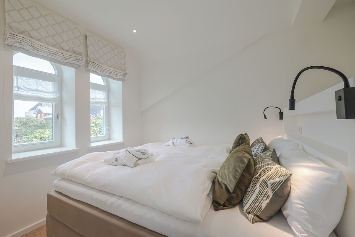 Quarto com cama, almofadas, janela com cortinas e luminária na parede.