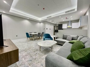Quarto de hotel moderno com piso de mármore, sofá, mesa de centro, televisão e área de jantar com cadeiras e mesa.