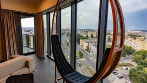 Quarto moderno com uma cadeira pendular curvada, um sofá e grandes janelas com vista para a cidade.