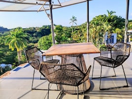 Um pátio externo com mesa de madeira e cadeiras de vime, rodeado por vegetação exuberante e uma vista desobstruída da paisagem.