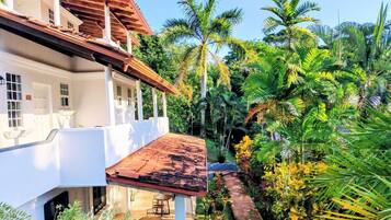 Um edifício branco com telhado de telhas vermelhas, um alpendre coberto com mesas e um jardim exuberante com palmeiras.