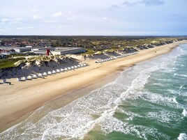 Uma praia com uma fileira de cabanas de praia brancas, um edifício grande com várias janelas e uma faixa de areia com ondas.