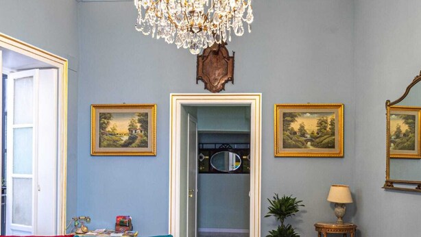 Um quarto espaçoso com um grande candelabro, um sofá azul, uma mesa de centro de vidro e quadros de paisagens nas paredes.