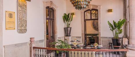 Um saguão de hotel com teto de madeira, um lustre decorativo em formato de folha, plantas em vasos, um balcão de recepção e uma placa com 'Casa Kloster'.