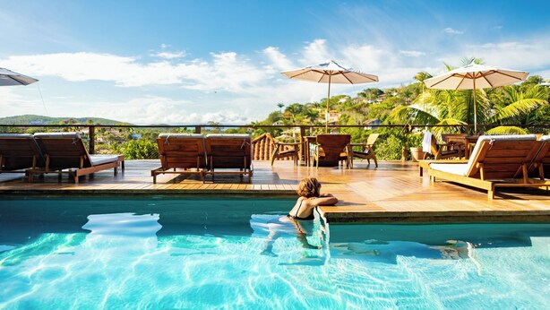 Uma área ao lado da piscina com espreguiçadeiras de madeira e guarda-sóis, cercada por vegetação exuberante e um céu claro.