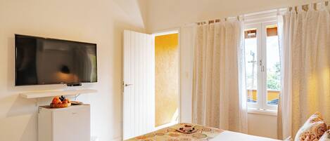 Quarto de hotel com uma cama, televisão fixada na parede, mini-geladeira e janela com cortinas.