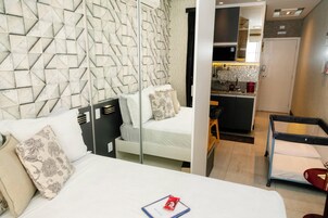 Quarto de hotel com design moderno, apresentando uma parede com padrão geométrico, uma cama branca com almofadas estampadas, uma pequena escrivaninha e uma cozinha compacta ao fundo.