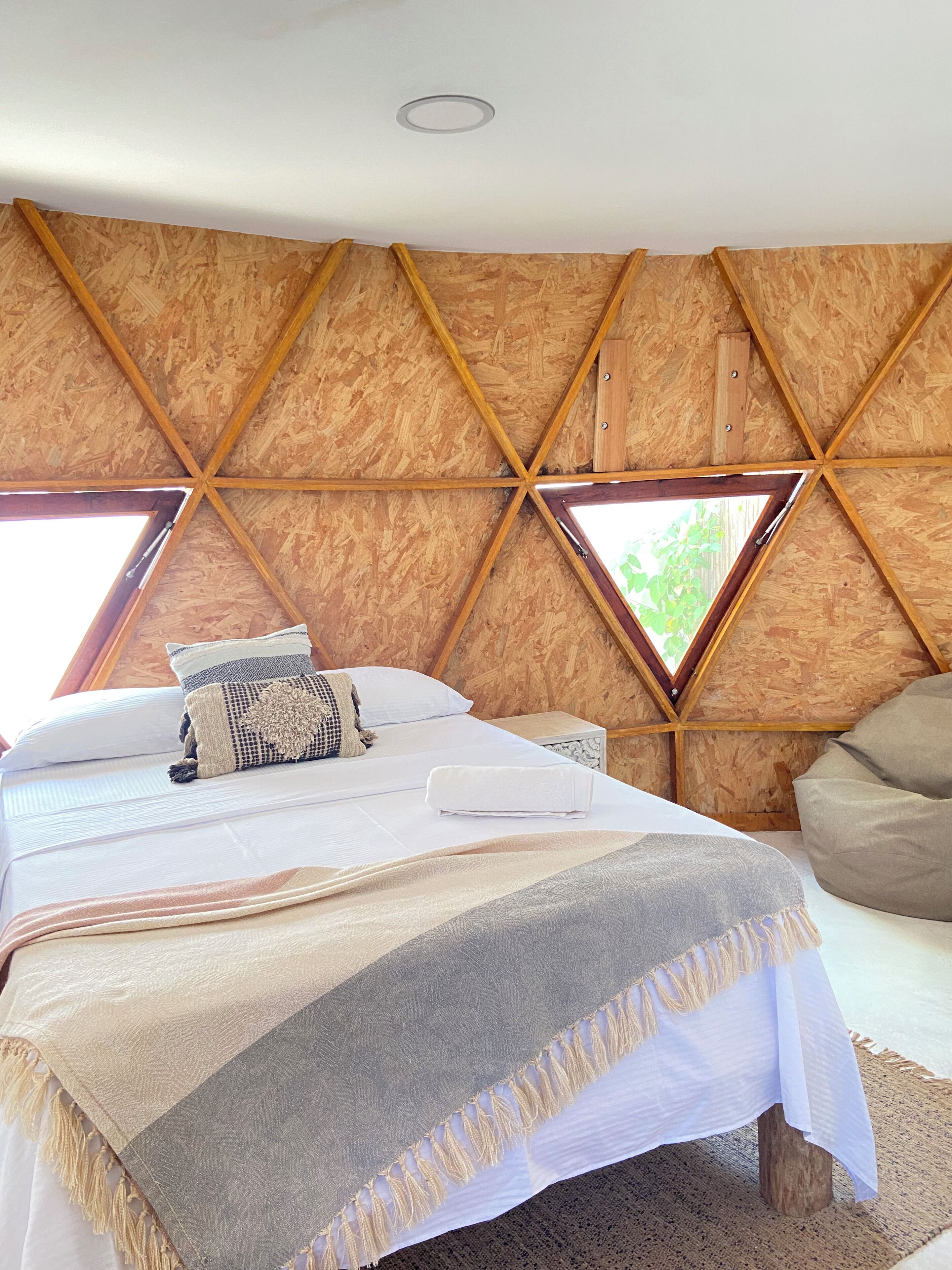 Quarto com teto de madeira, cama com colchão branco e manta bege, um travesseiro e uma mesinha de cabeceira.
