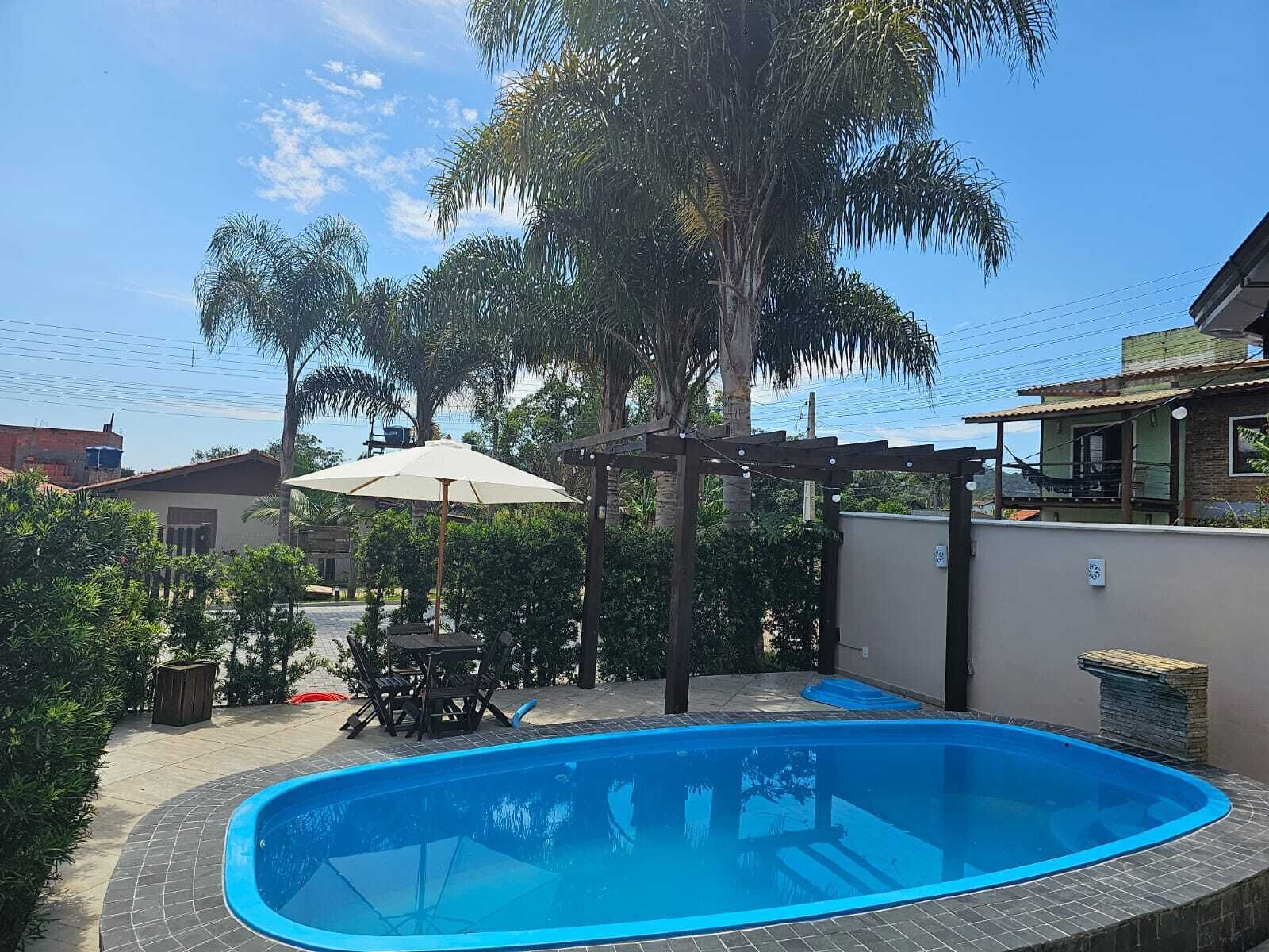 Piscina com borda azulejada, rodeada por um espaço de pátio com mesa e cadeiras, e um pergolado com guarda-sol.