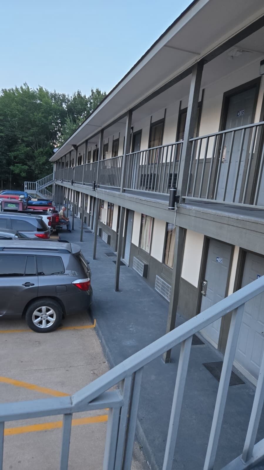 Um motel com vários andares, uma área de estacionamento, um homem sentado num banco e um céu claro.
