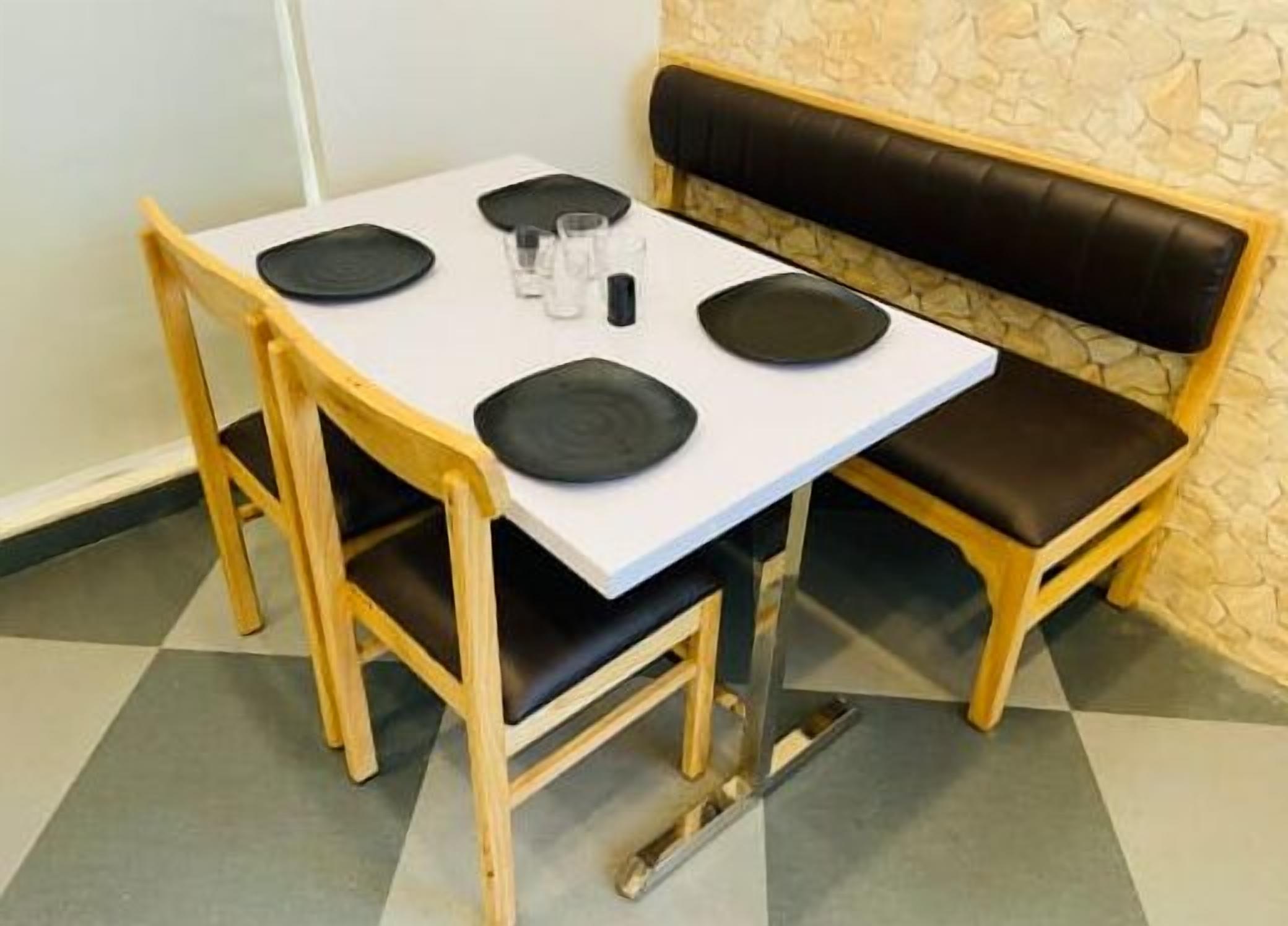 Uma mesa de jantar com quatro pratos pretos, dois copos e uma garrafa pequena. Há duas cadeiras de madeira com almofadas pretas e um banco com almofada preta. A mesa é branca, e o chão tem padrão xadrez.