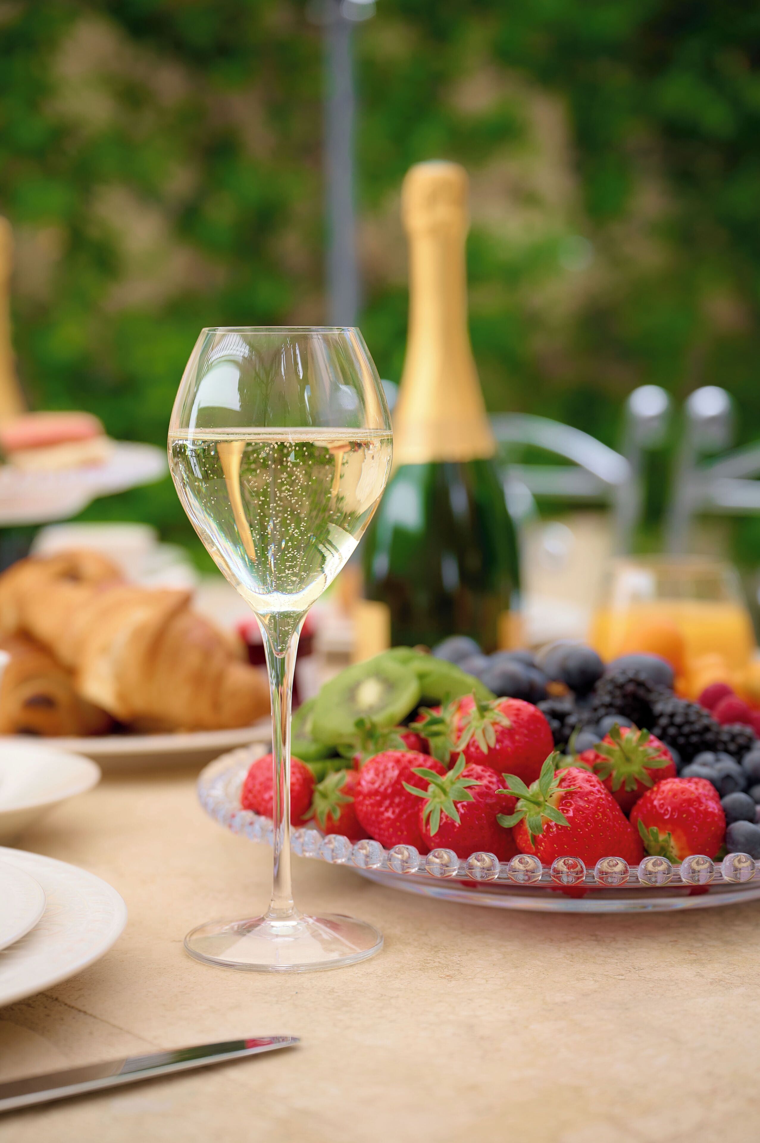 Um copo de vinho branco, uma garrafa de champanhe e um prato com frutas vermelhas frescas e doces numa mesa.