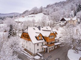 Uma casa de madeira de vários andares com telhado inclinado, rodeada por árvores cobertas de neve e uma paisagem acidentada.