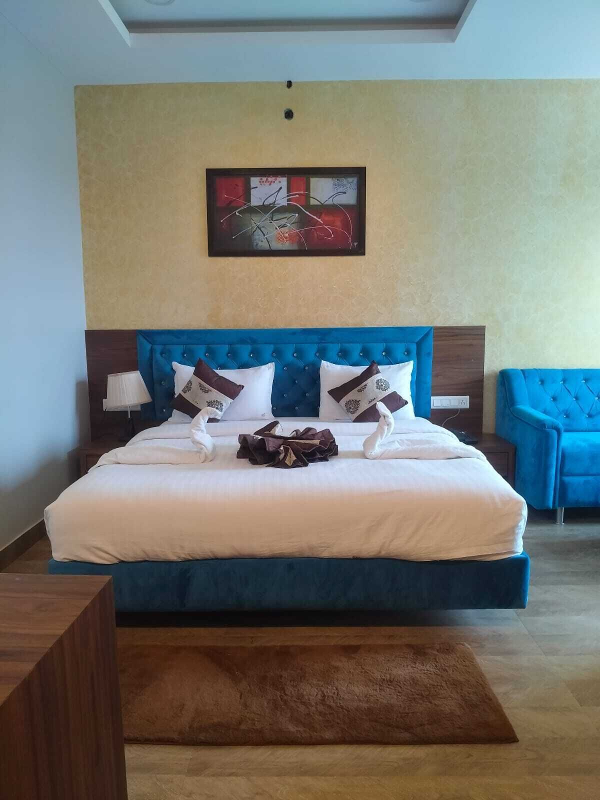 Quarto de hotel com uma cama grande, uma poltrona azul estofada, um quadro na parede e um tapete castanho.