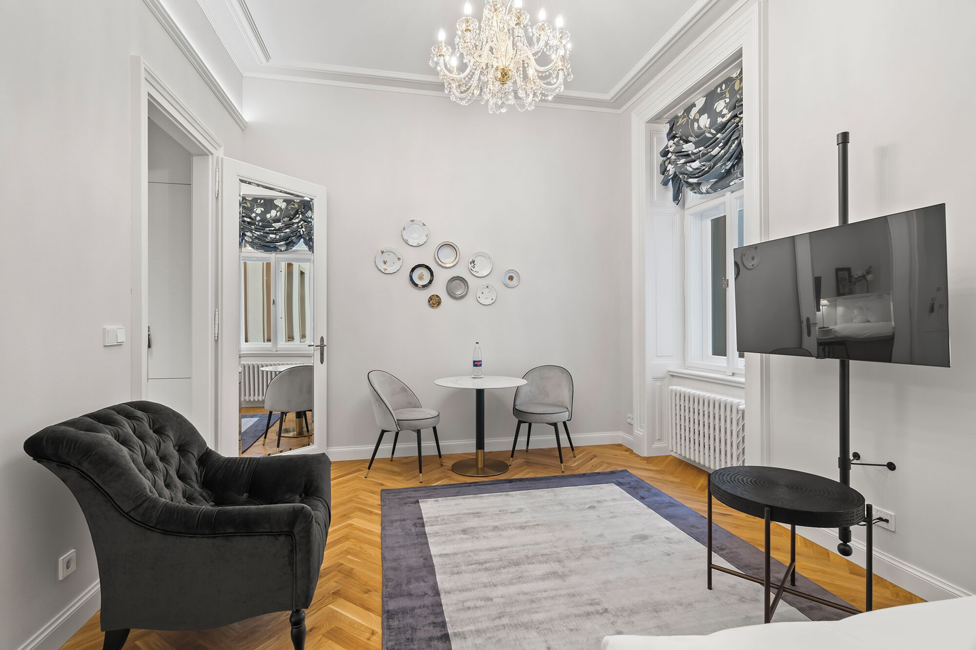 Uma sala de estar moderna com uma poltrona de veludo preta, uma mesa de centro redonda e uma TV sobre um suporte. Há duas cadeiras ao redor de uma mesa pequena, e um candelabro pendurado no teto.