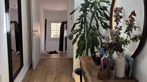 Um corredor com piso de madeira, um espelho grande, um vaso de planta, uma mesa com revistas e um claraboia.