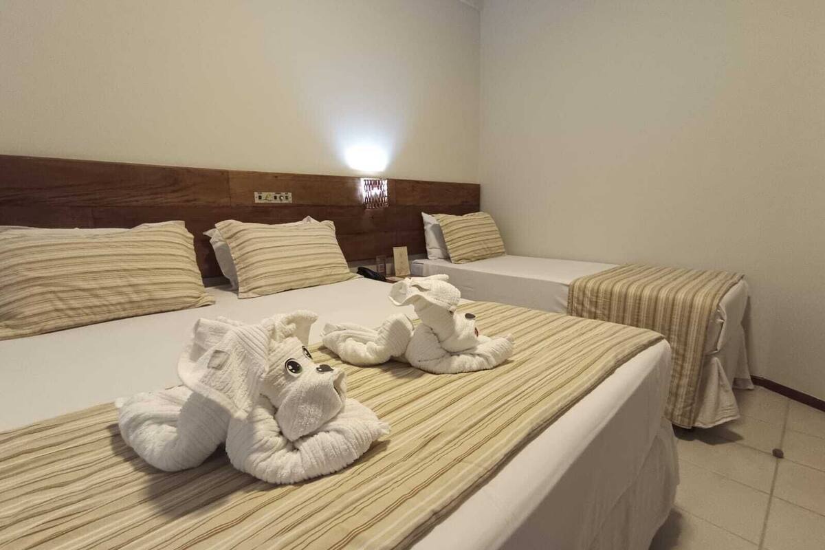 Quarto de hotel com duas camas, cada uma com roupa de cama listrada e cabeceira de madeira. Há toalhas em formato de elefante sobre as camas.