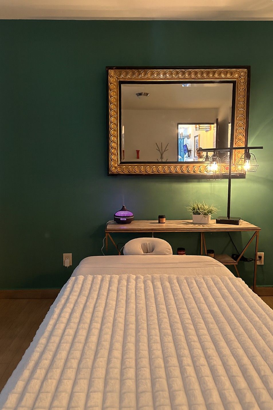 Cama bem arrumada com colcha branca, mesa de cabeceira com abajur, espelho e uma pequena planta em vaso.