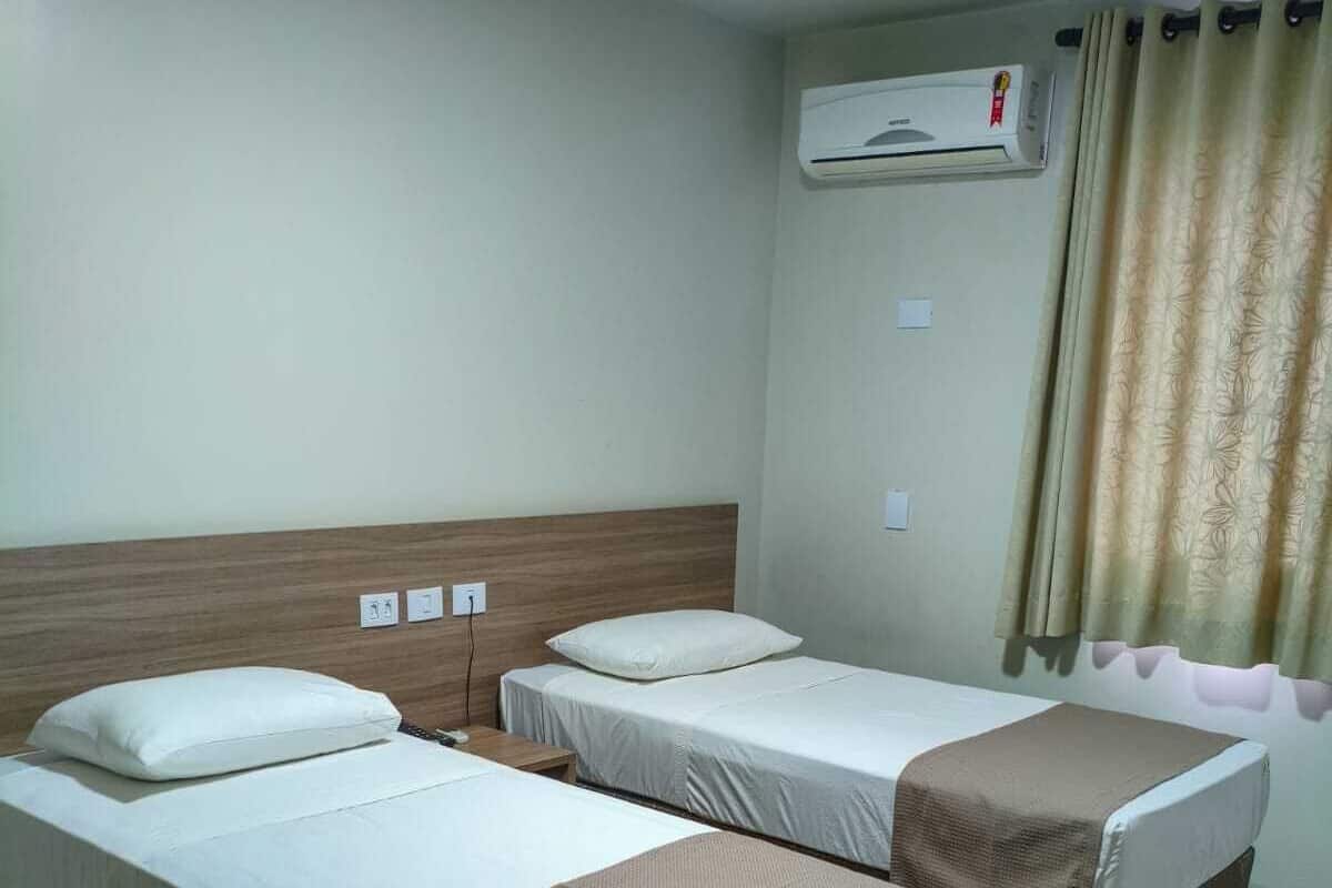 Quarto de hotel com duas camas de solteiro, cabeceira de madeira, ar condicionado e cortina.