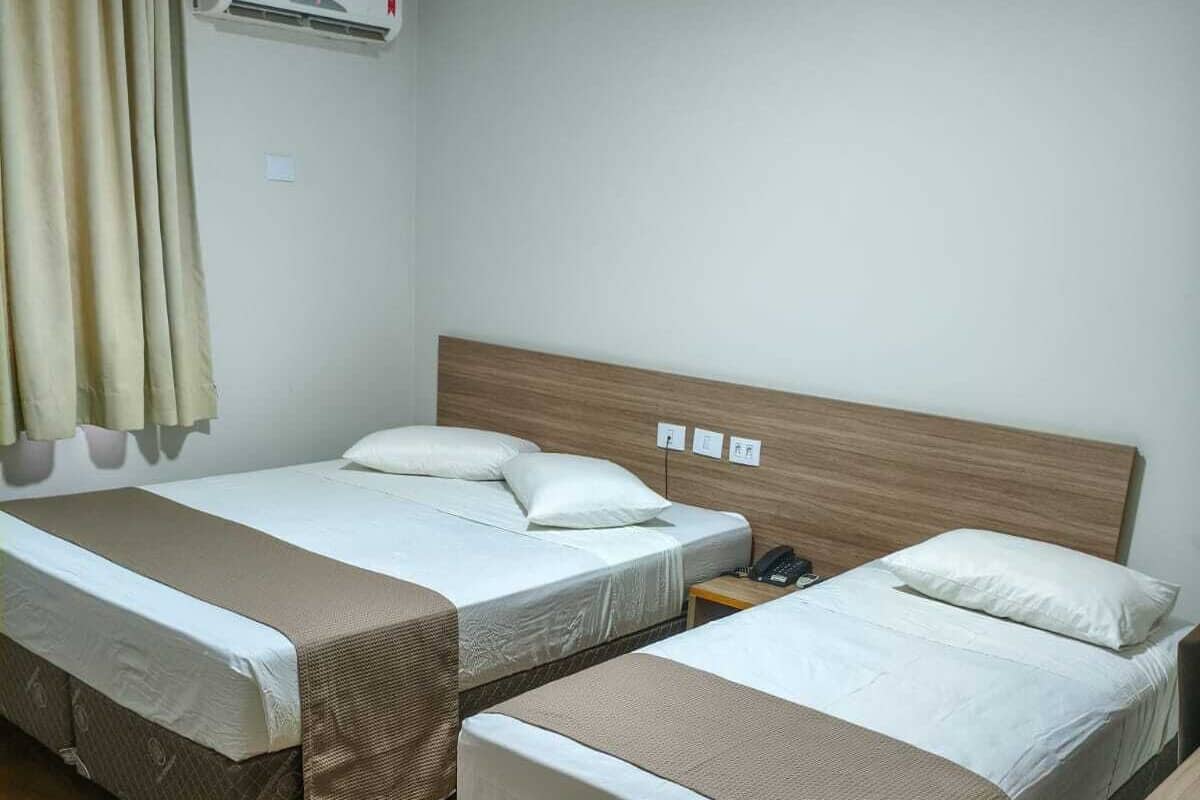 Quarto com duas camas de solteiro, cabeceira de madeira, ar condicionado fixado na parede e cortina.