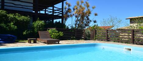 Piscina com deck de madeira, área de estar coberta e jardim cercado.