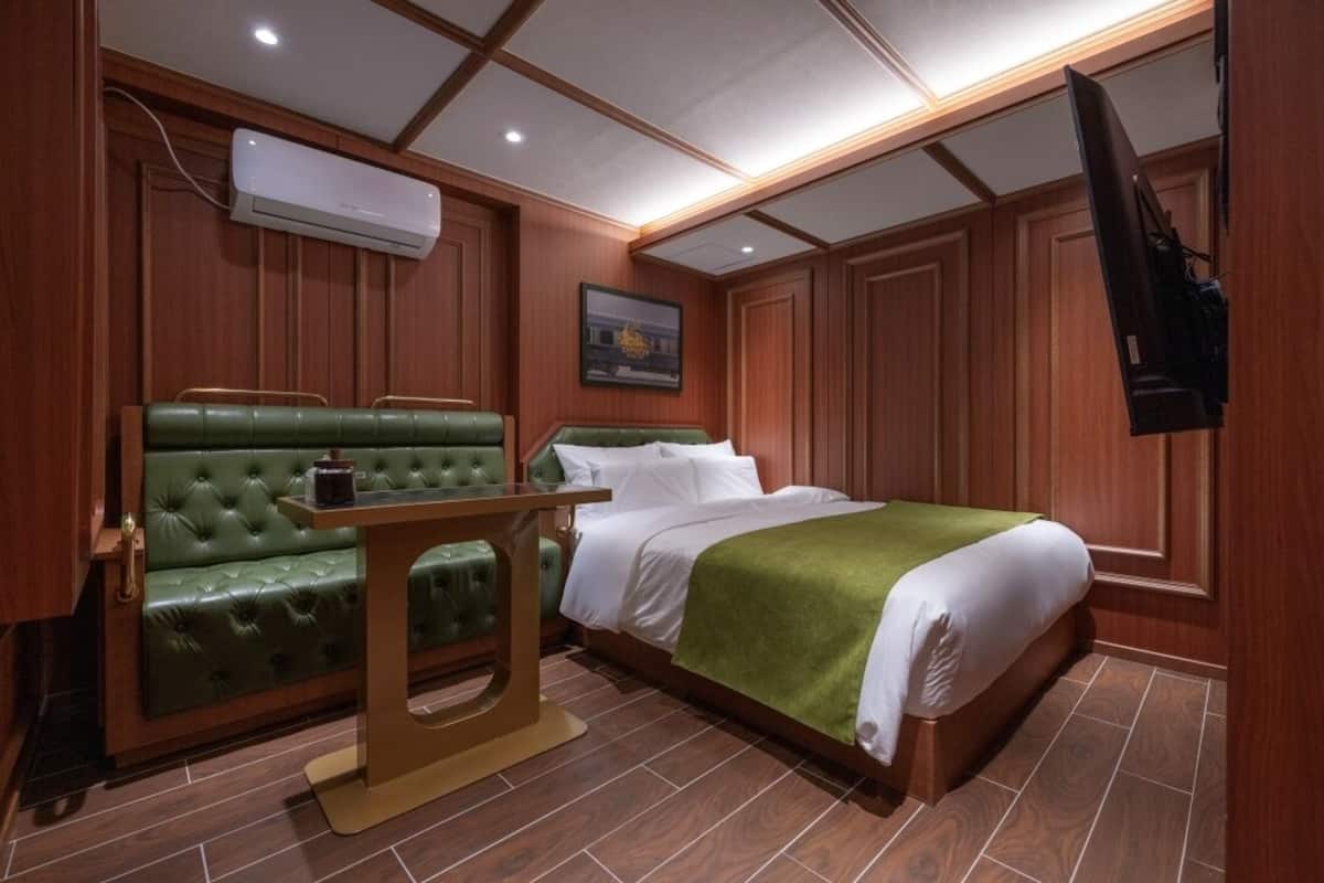 Quarto de hotel com sofá de couro verde, cama de madeira com manta verde, mesa pequena e ar condicionado na parede.
