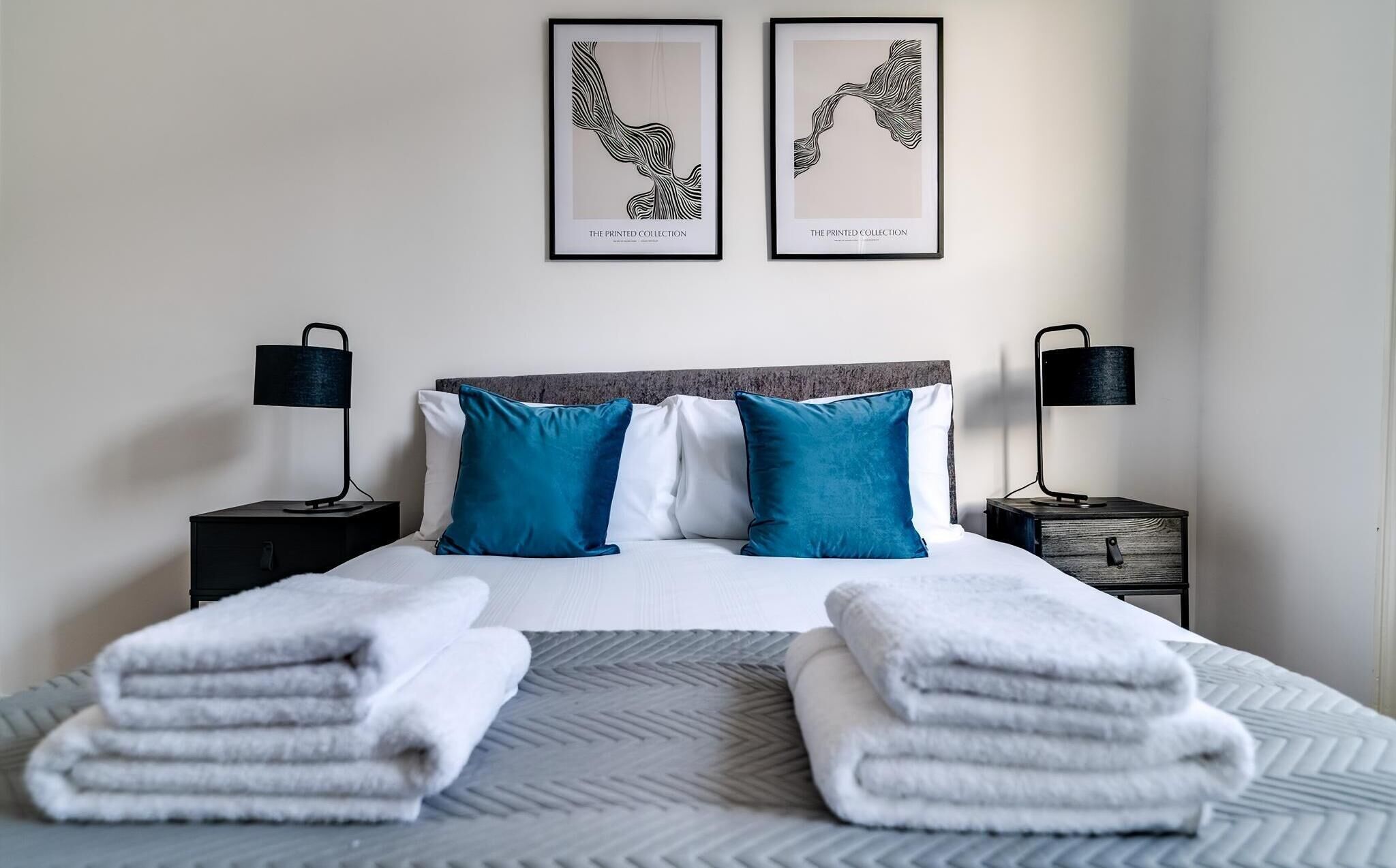 Cama bem arrumada com duas almofadas, um edredom cinza e branco estampado, e duas toalhas brancas sobre a cama. Há duas mesinhas de cabeceira pretas com abajures, e duas obras de arte emolduradas acima da cama.