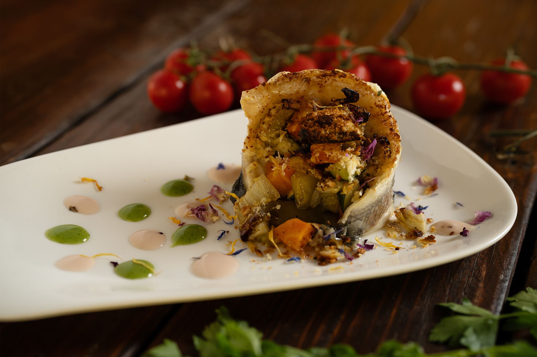 Um prato com exterior crocante, possivelmente um vegetal ou carne recheado, sobre um prato branco com flores comestíveis verdes e rosa espalhadas.