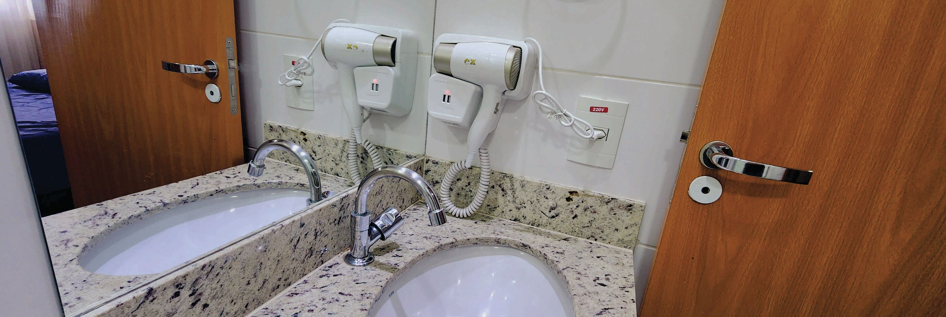 Casa de banho com duas pias, um espelho e dois secadores de cabelo fixados na parede.
