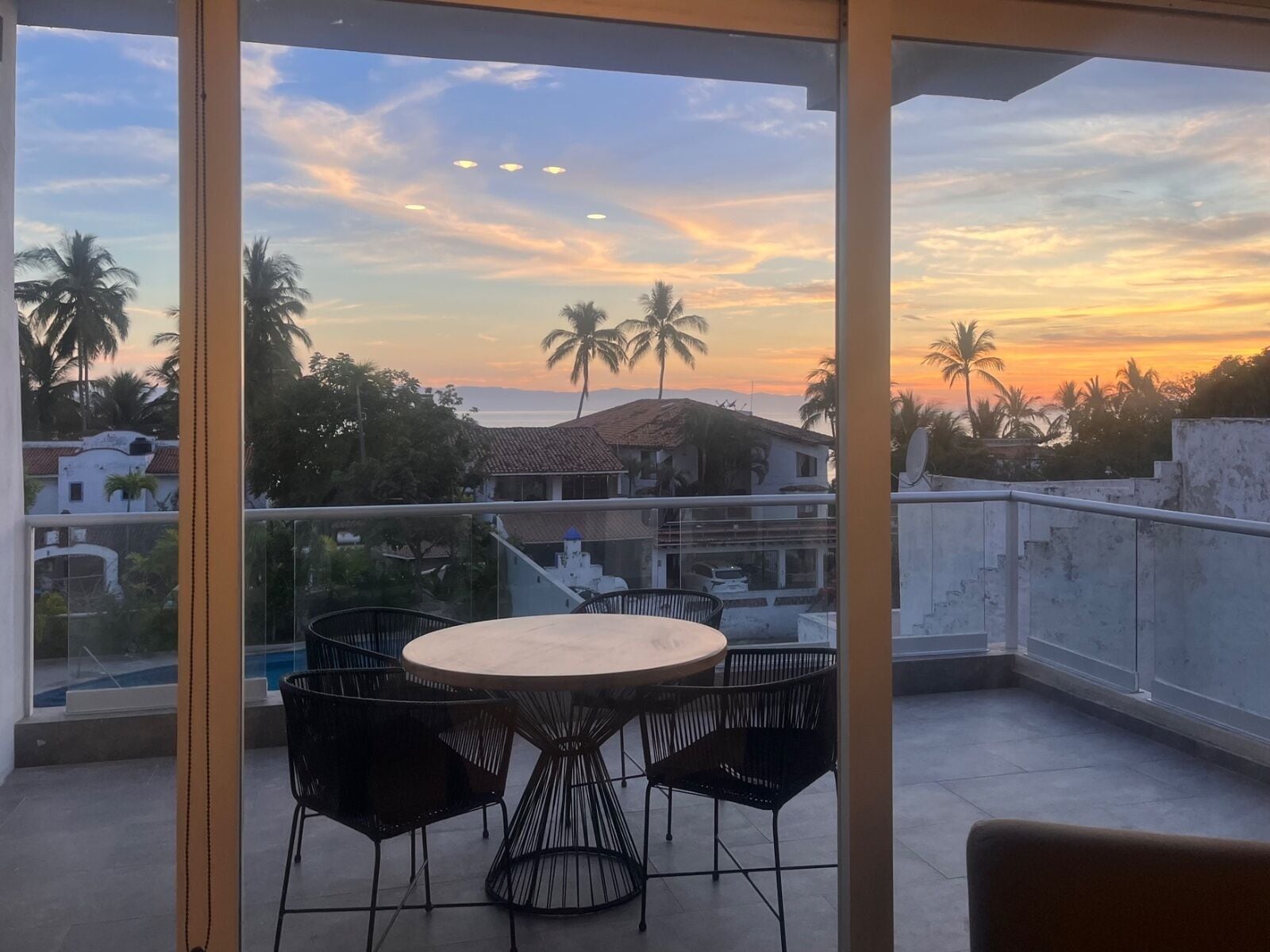 Uma varanda com uma mesa redonda e cadeiras, com vista para o pôr do sol, palmeiras e edifícios ao fundo.