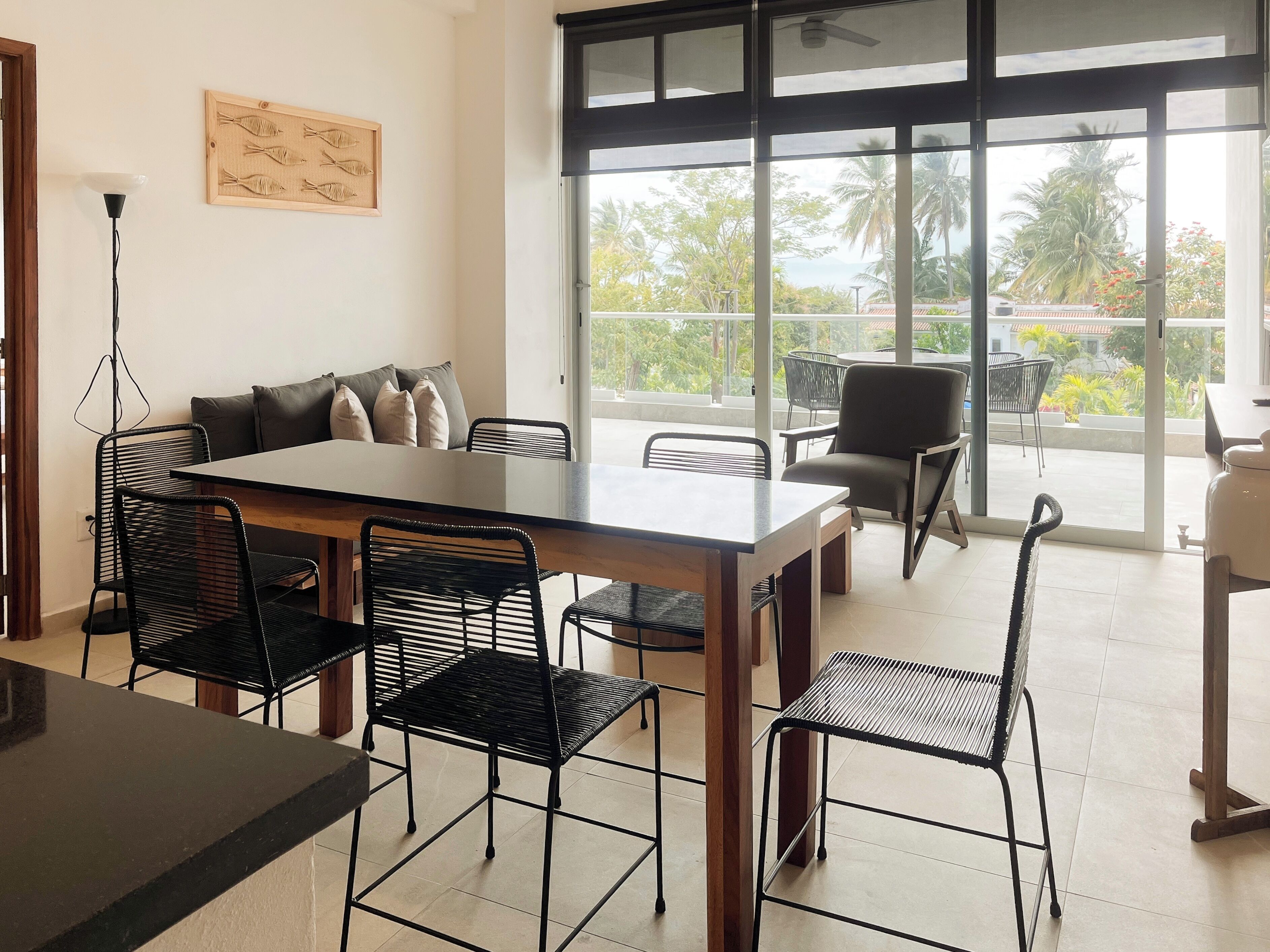 Uma área de refeições moderna com mesa de madeira e cadeiras de metal preto, um sofá com almofadas e vista para palmeiras através de grandes janelas.