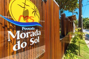 Uma placa de madeira com o texto 'Pousada Morada do Sol' e um logotipo com um sol e palmeiras.