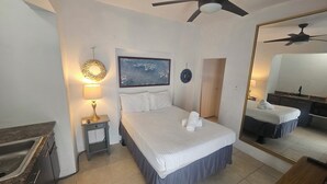 Quarto de hotel com cama, escrivaninha, espelho, ventoinha no teto e um quadro na parede.