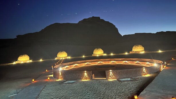 Uma grande estrutura em forma de cúpula iluminada, com um caminho circular que a leva, ao fundo, uma paisagem montanhosa.