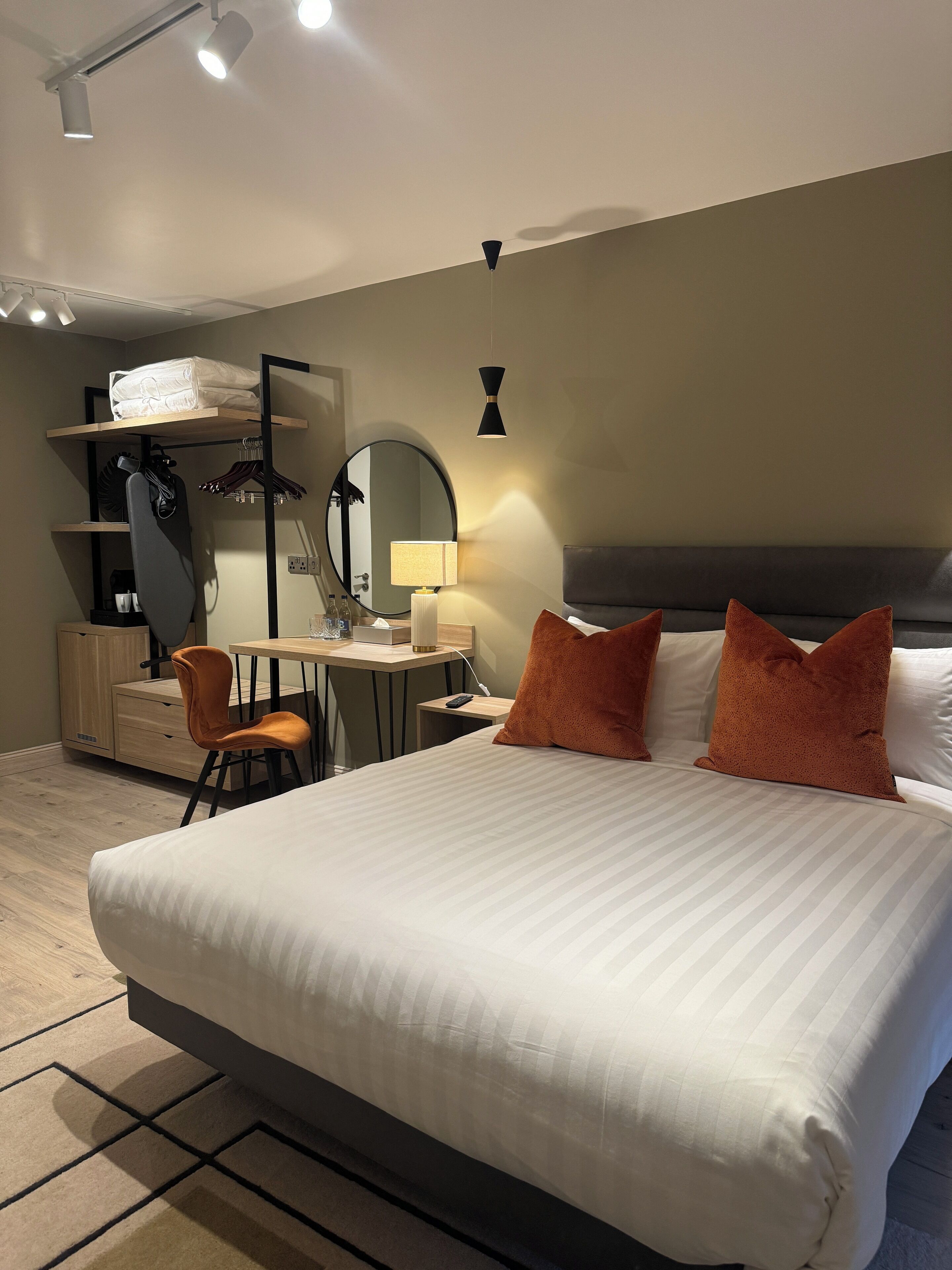 Quarto de hotel bem arrumado, com uma cama grande, uma escrivaninha com cadeira, guarda-roupa e um espelho.