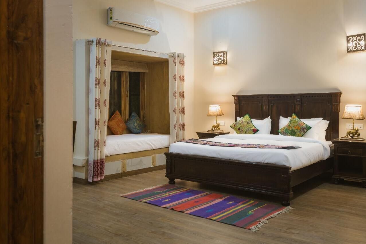 Quarto com cama grande, duas mesas de cabeceira com luminárias, tapete colorido e janela com cortinas.