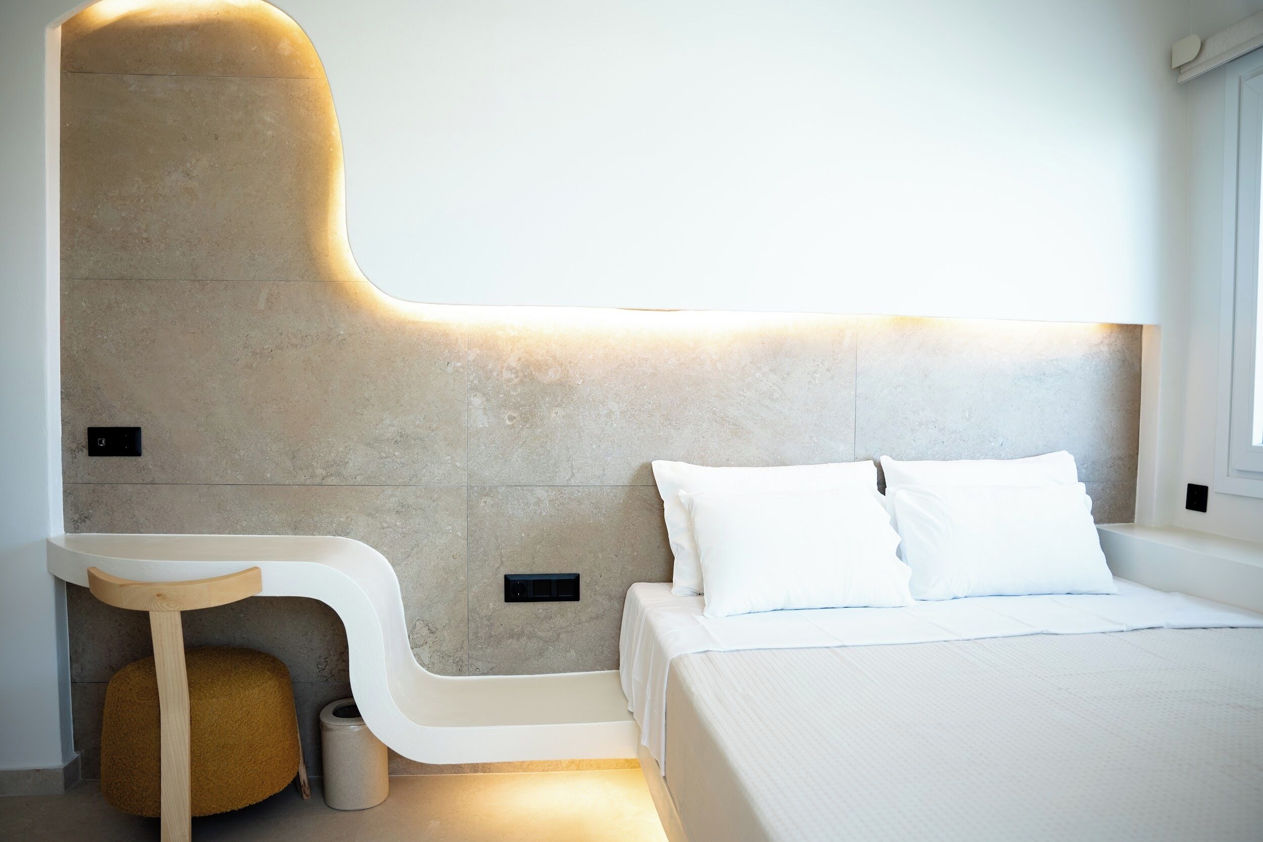 Quarto moderno com cabeceira curva, mesa de cabeceira e um banquinho redondo.