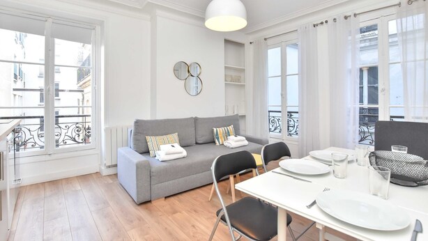 Uma sala de estar moderna com um sofá cinzento, uma mesa de jantar posta com pratos e copos, e uma luminária pendente branca.