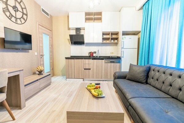 Uma sala de estar moderna com um sofá, uma mesa de centro com frutas, uma televisão e uma cozinha ao fundo.