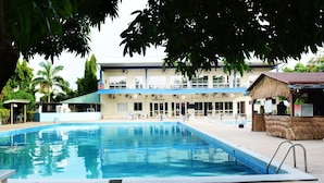 Uma piscina com água azul cristalina, rodeada por um deck de concreto. Há espreguiçadeiras e uma pequena construção com telhado de palha.