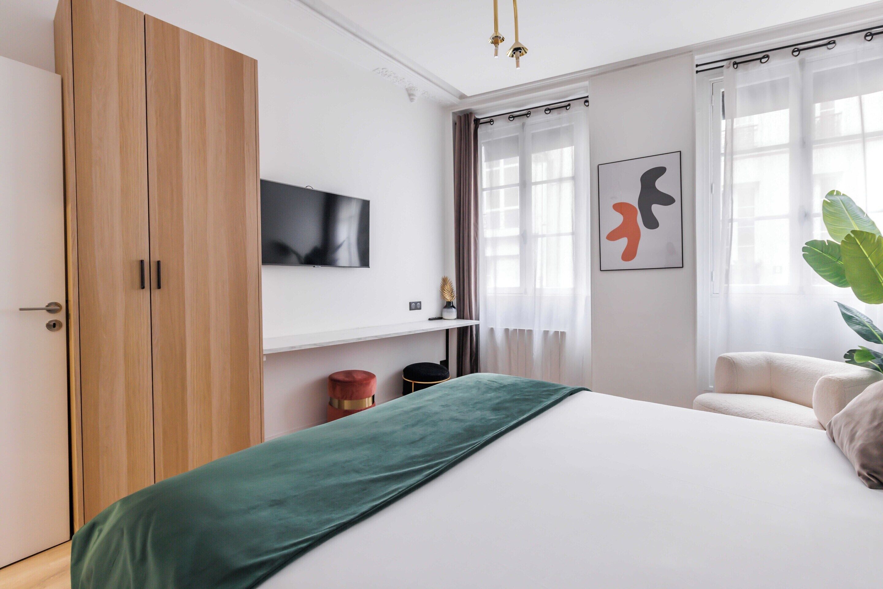 Quarto com cama, televisão na parede, planta e janela com cortinas.