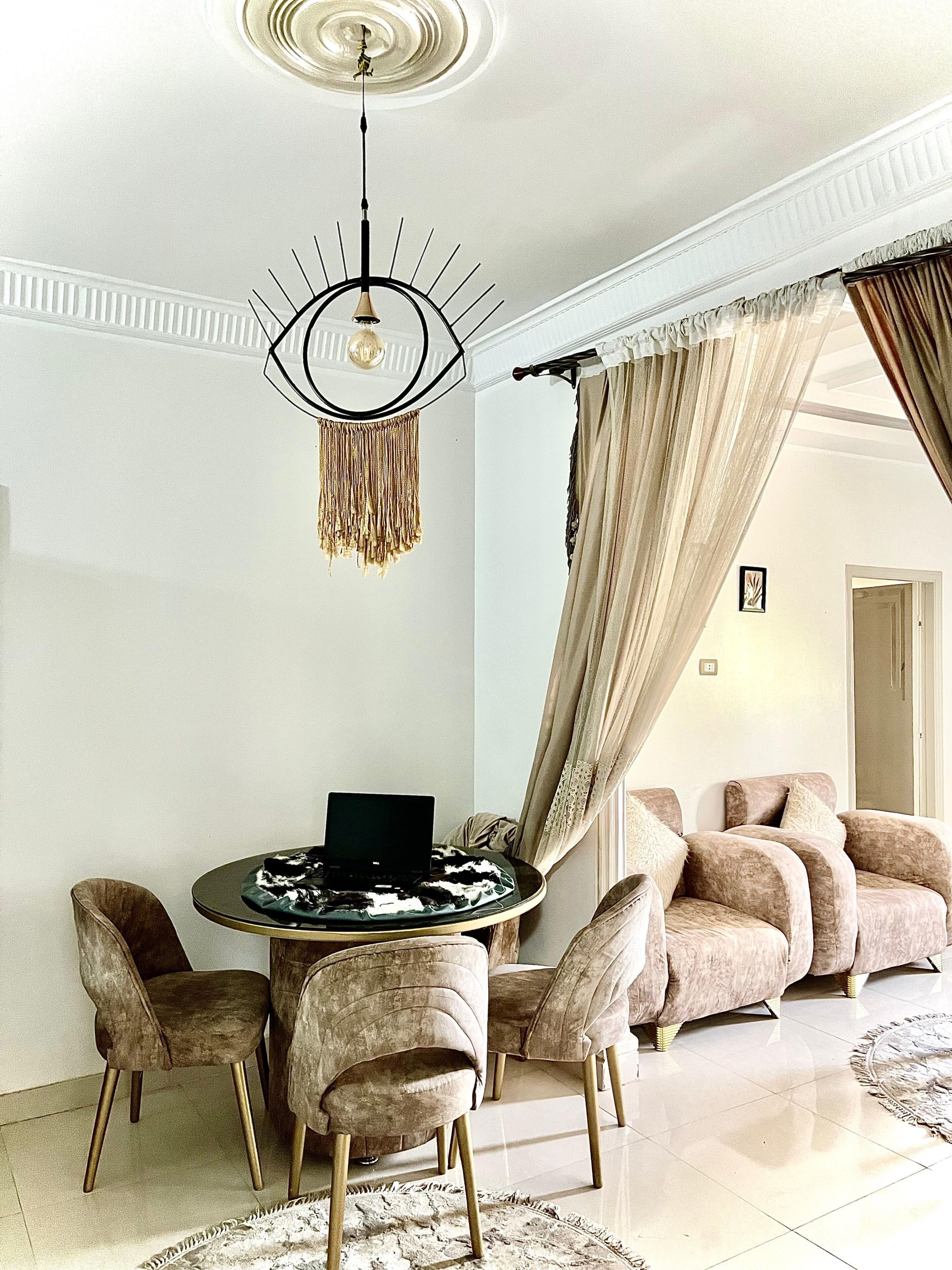 Uma sala de estar moderna com uma mesa de centro redonda, um laptop e um candelabro. Há poltronas confortáveis e um sofá com cortinas.