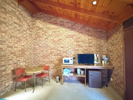 Um quarto pequeno com parede de tijolos, teto de madeira, uma mesa redonda com duas cadeiras vermelhas, uma televisão e uma geladeira pequena.