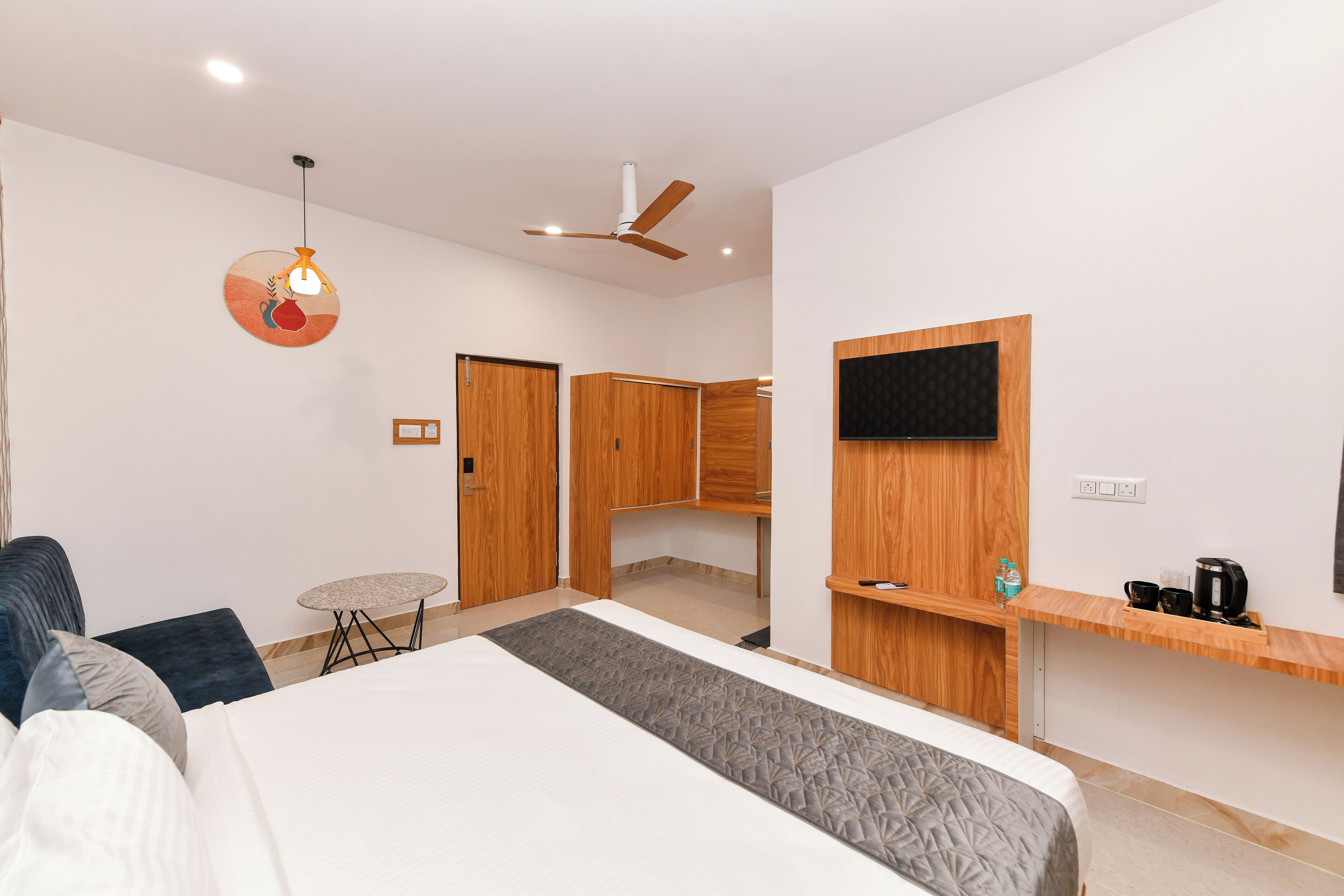 Quarto de hotel moderno com uma cama grande, uma mesa redonda pequena, um ventilador de teto e uma televisão na parede.