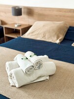 Cama bem arrumada com colcha azul, almofada bege e duas toalhas brancas enroladas com etiquetas verdes sobre um tapete bege texturizado.