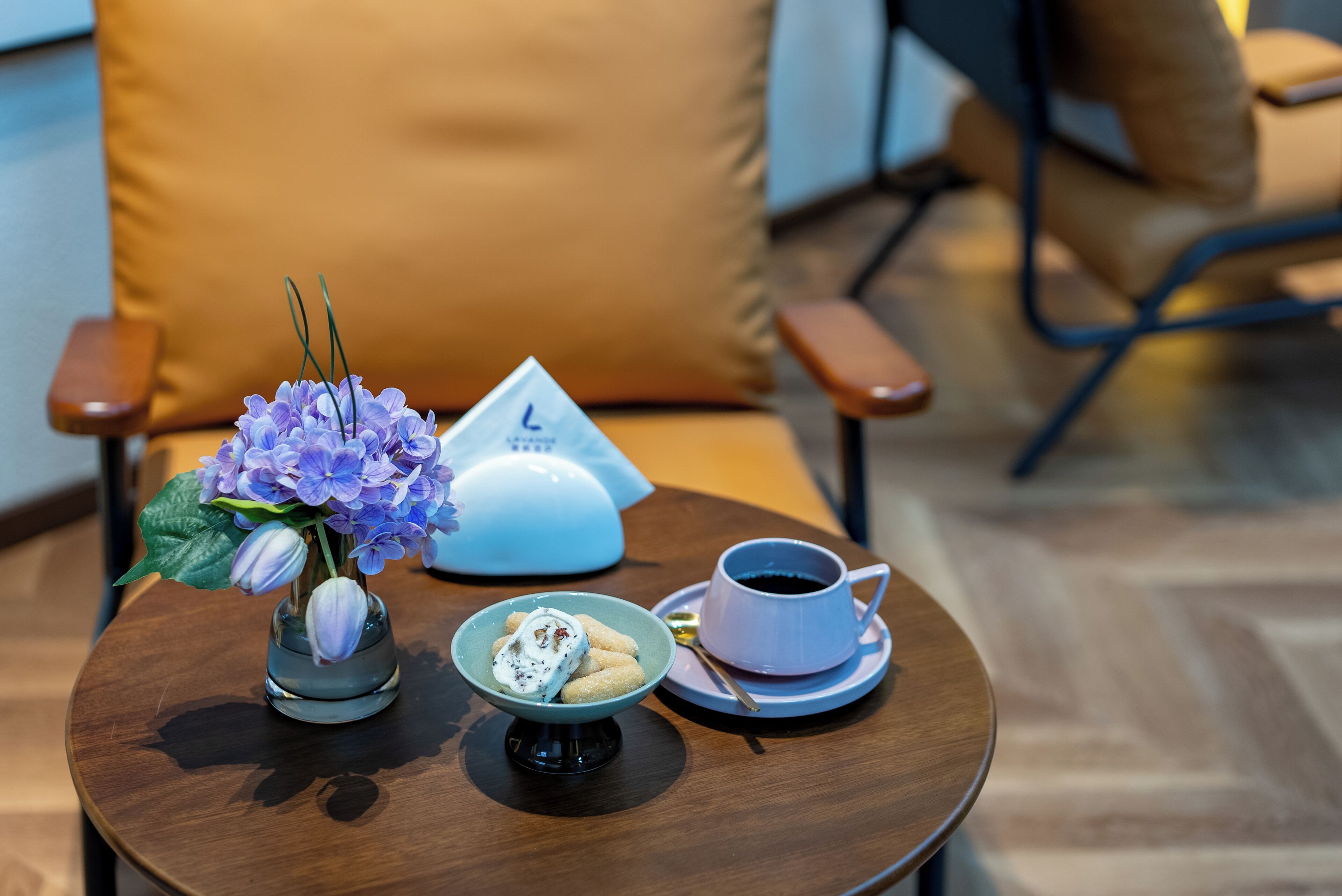 Uma mesa redonda de madeira com uma chaleira branca, uma chávena de café, uma tigela de bolachas e um vaso de flores roxas.