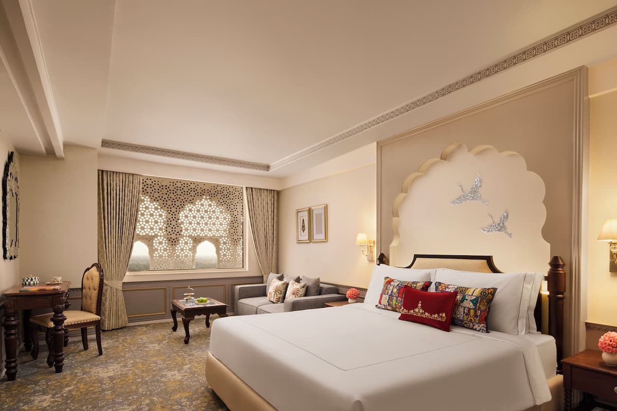 Quarto de hotel com uma cama grande, uma escrivaninha, uma cadeira, uma mesinha e uma janela com cortinas.