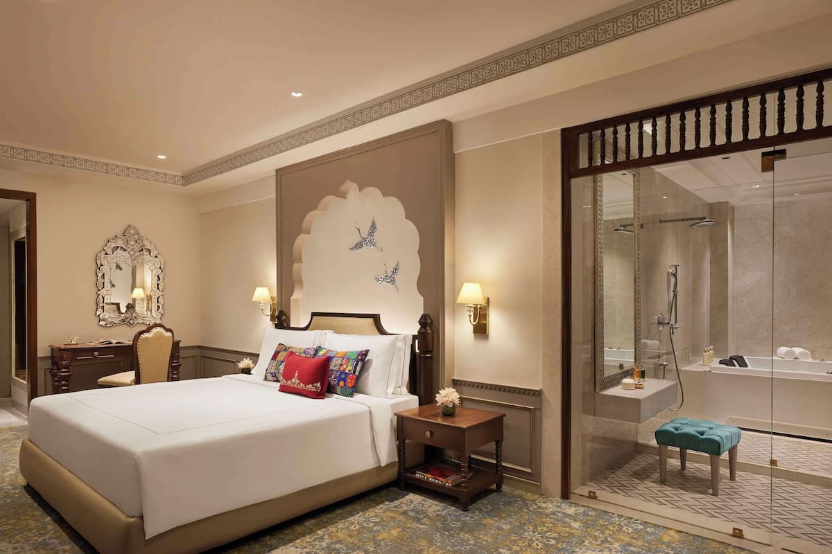 Quarto de hotel com uma cama grande, mesa de cabeceira, espelho, área de chuveiro e divisória de vidro.